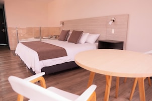 Quarto de hotel moderno com uma cama grande, mesas de cabeceira, uma mesa de madeira redonda e uma cadeira branca.