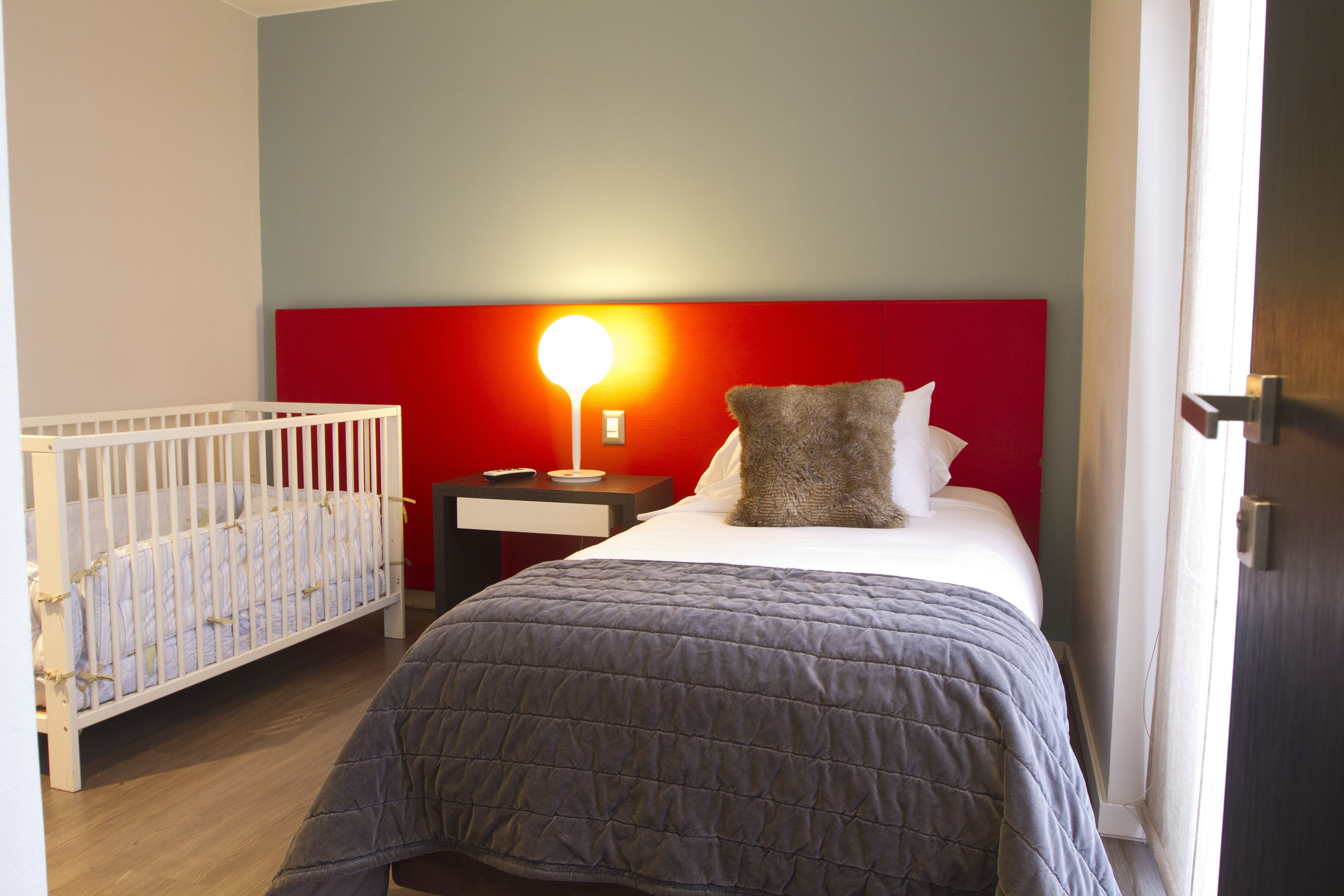 Quarto com berço, mesa de cabeceira com abajur, e cama com travesseiros e cobertor.
