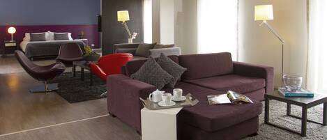 Quarto de hotel moderno com um sofá roxo, uma cadeira vermelha, uma pequena mesa lateral branca e um abajur.