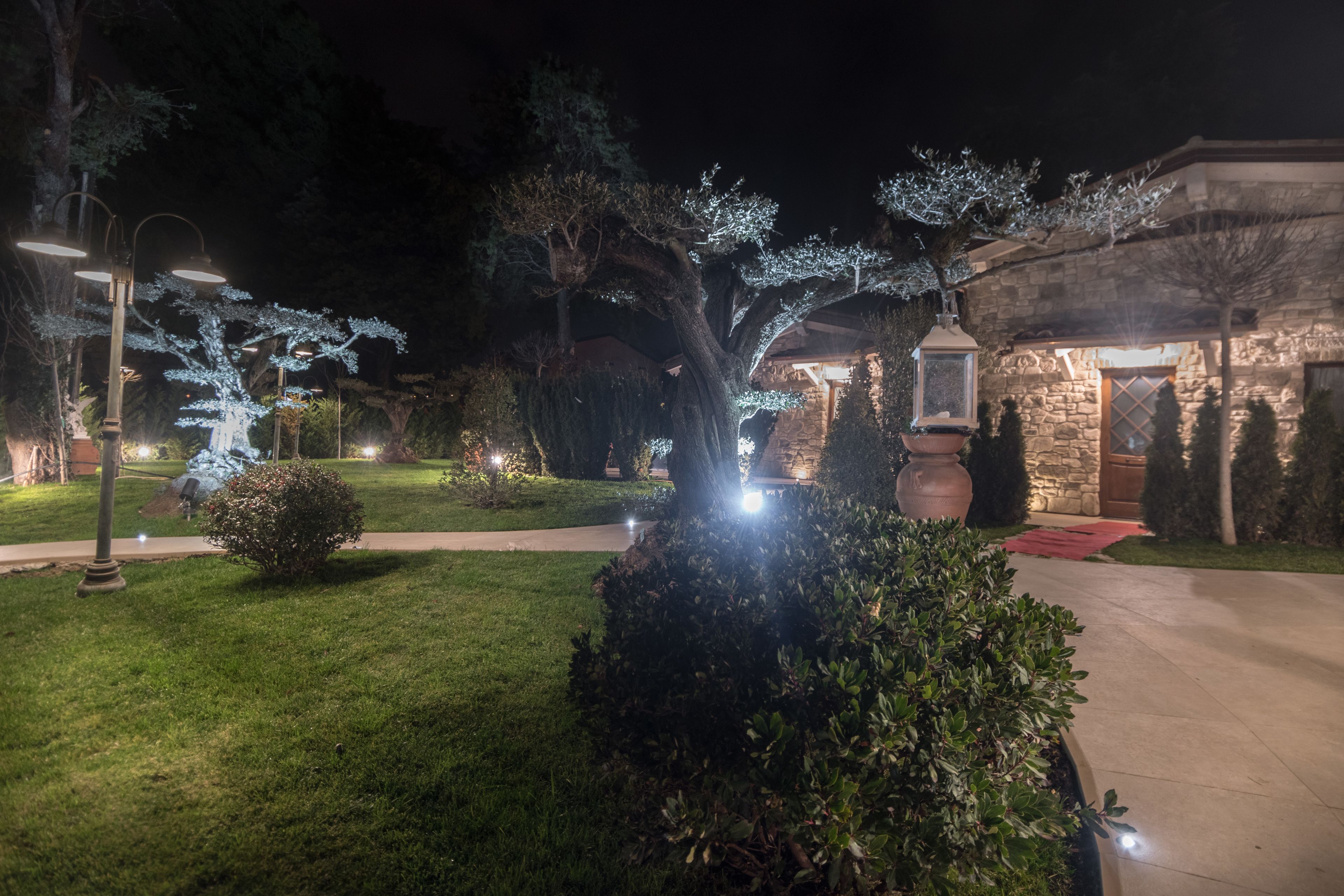 Um pátio bem iluminado com um edifício de pedra, um caminho e diversas árvores e arbustos.