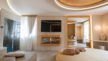 Quarto de hotel moderno com uma cama grande, lareira, televisão e chuveiro em vidro.
