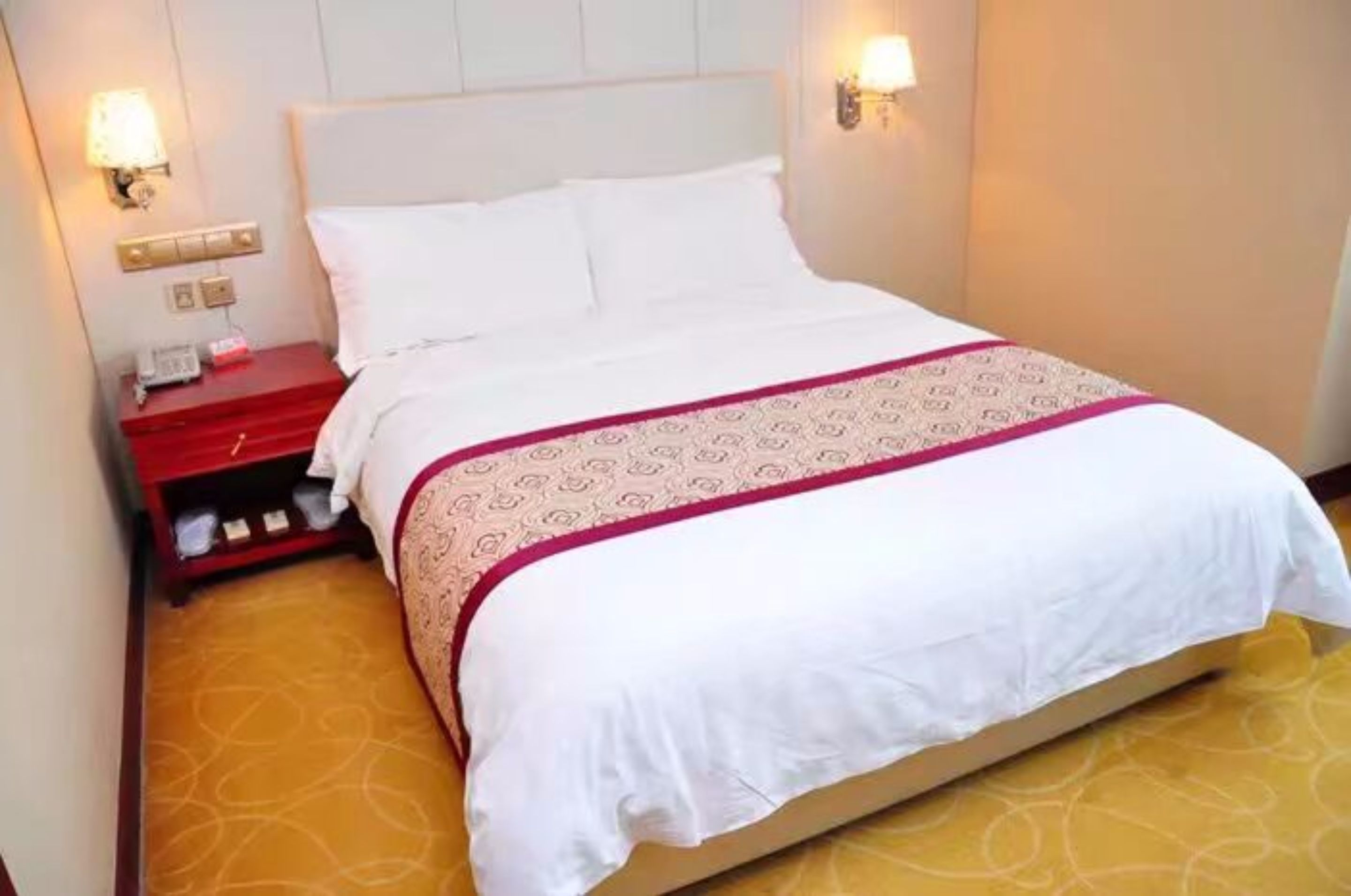 Quarto de hotel com uma cama, mesa de cabeceira e luminária fixada na parede.