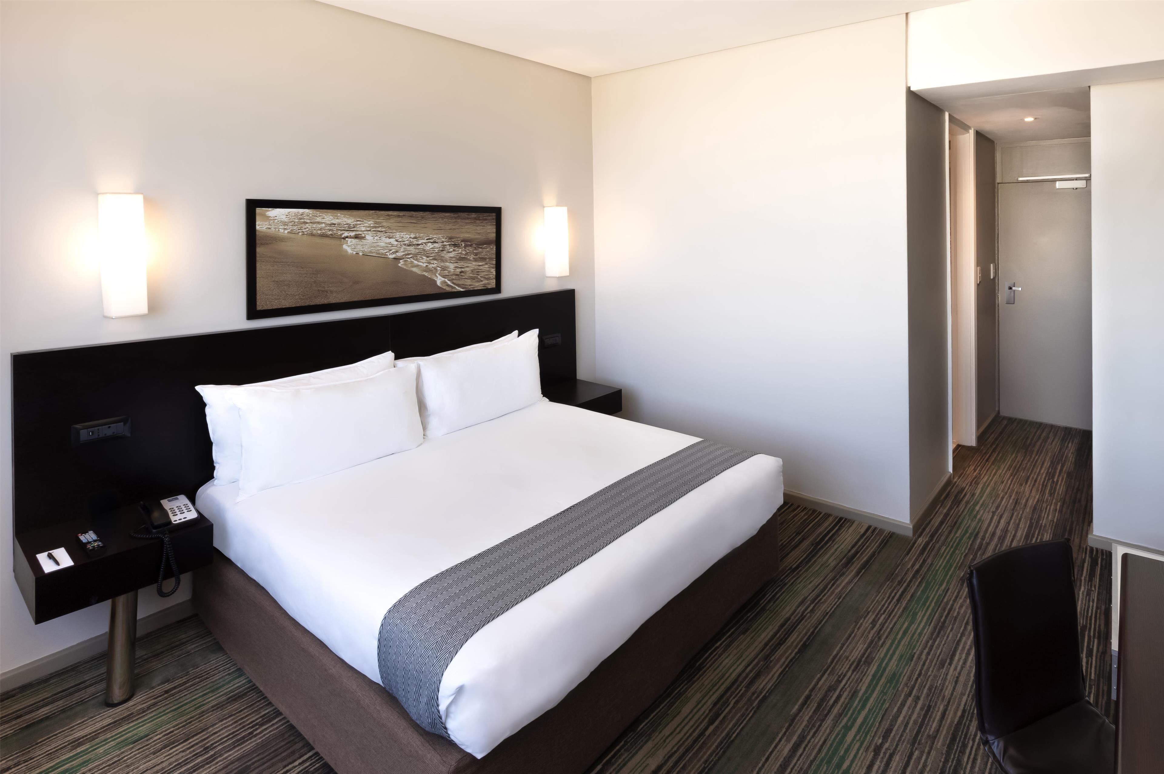 Quarto de hotel com uma cama grande, cabeceira, mesas de cabeceira, telefone e um quadro na parede.