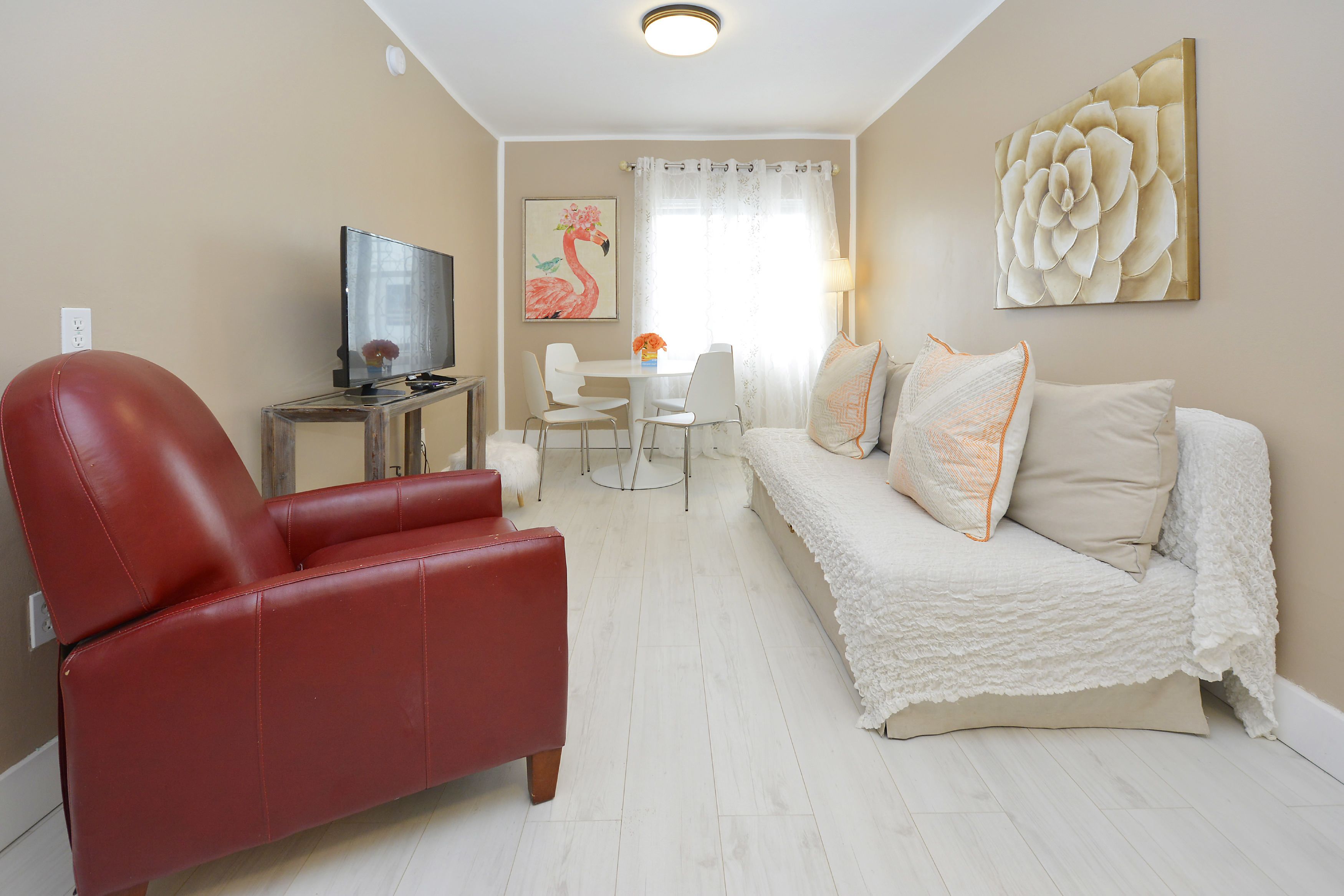 Uma sala de estar com uma poltrona de couro vermelho, um sofá branco, uma mesa de jantar com cadeiras, uma TV e obras de arte nas paredes.