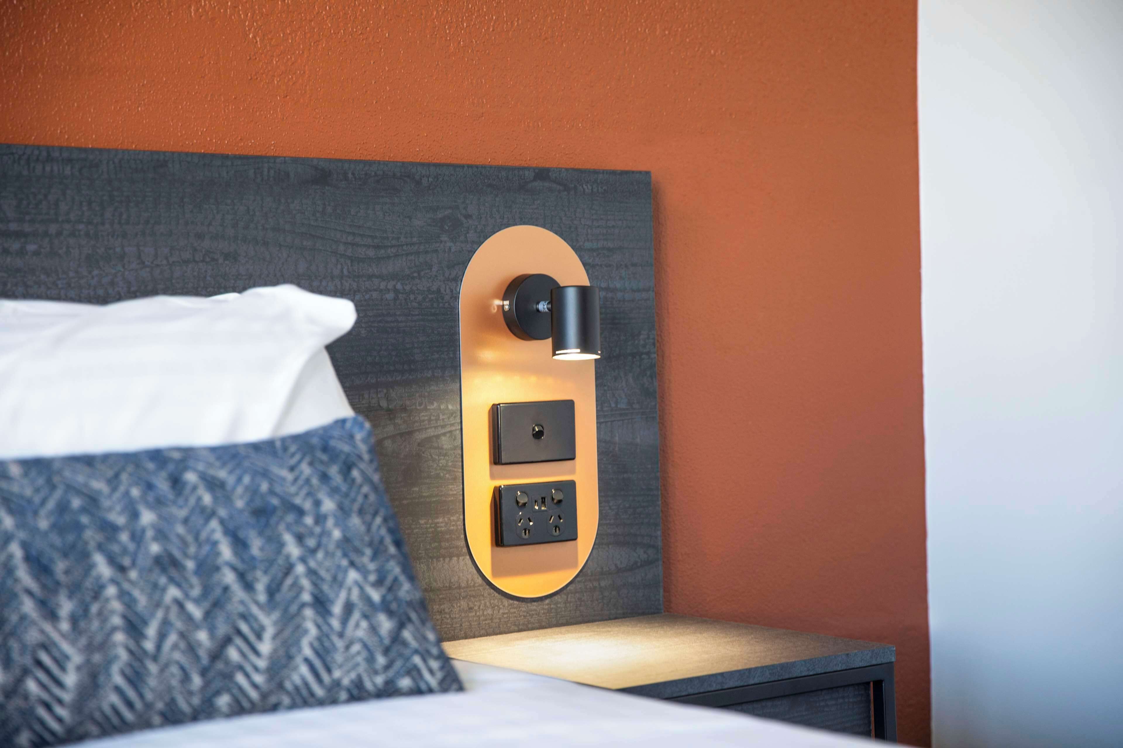 Quarto de hotel moderno com cabeceira de madeira, mesa de cabeceira e painel de controle fixado na parede.