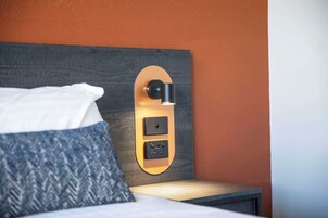 Quarto de hotel moderno com cabeceira de madeira, mesa de cabeceira e painel de controle fixado na parede.