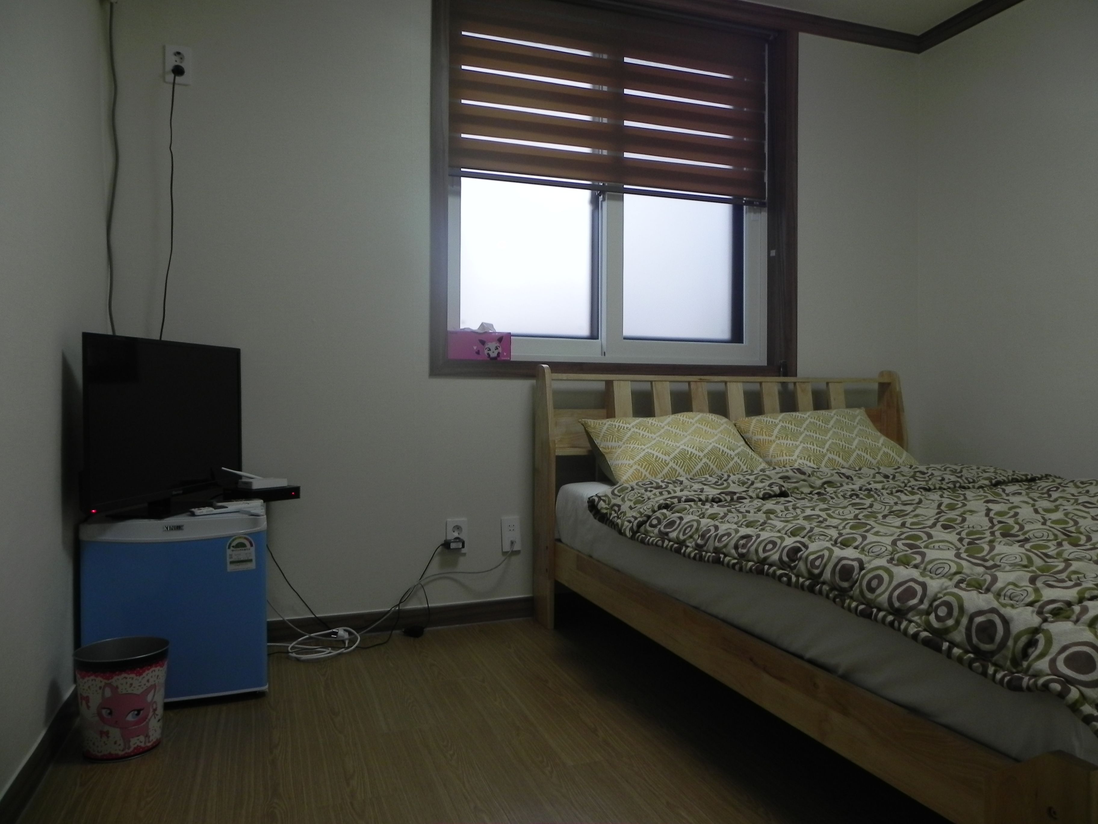 Quarto com cama, televisão sobre um suporte, armário azul pequeno, janela com persianas e tomada na parede.