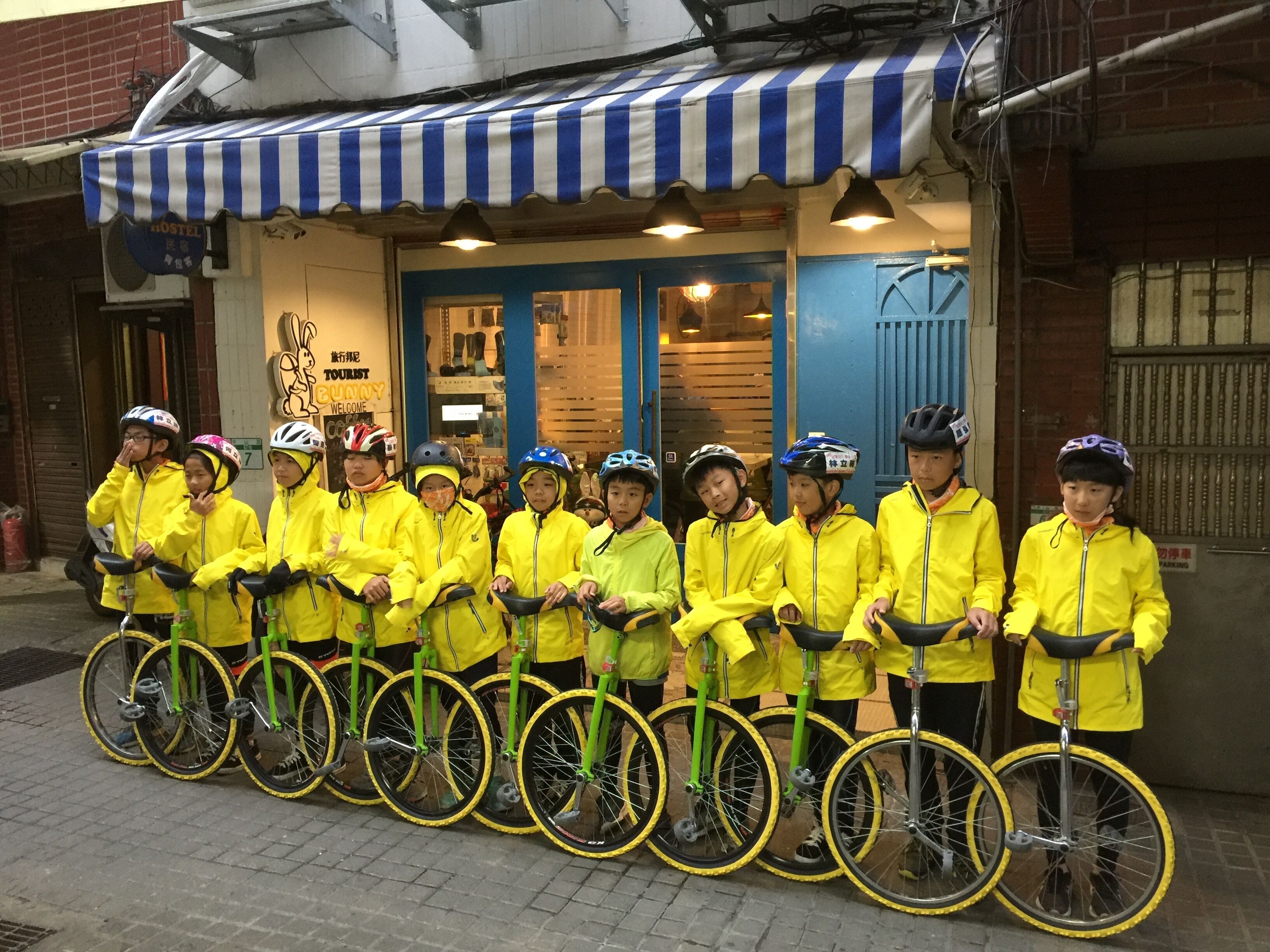 Um grupo de crianças de jaquetas amarelas em bicicletas em frente a um edifício com marquise azul e branca.