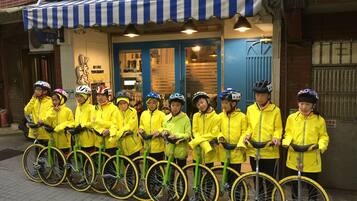 Um grupo de crianças de jaquetas amarelas em bicicletas em frente a um edifício com marquise azul e branca.