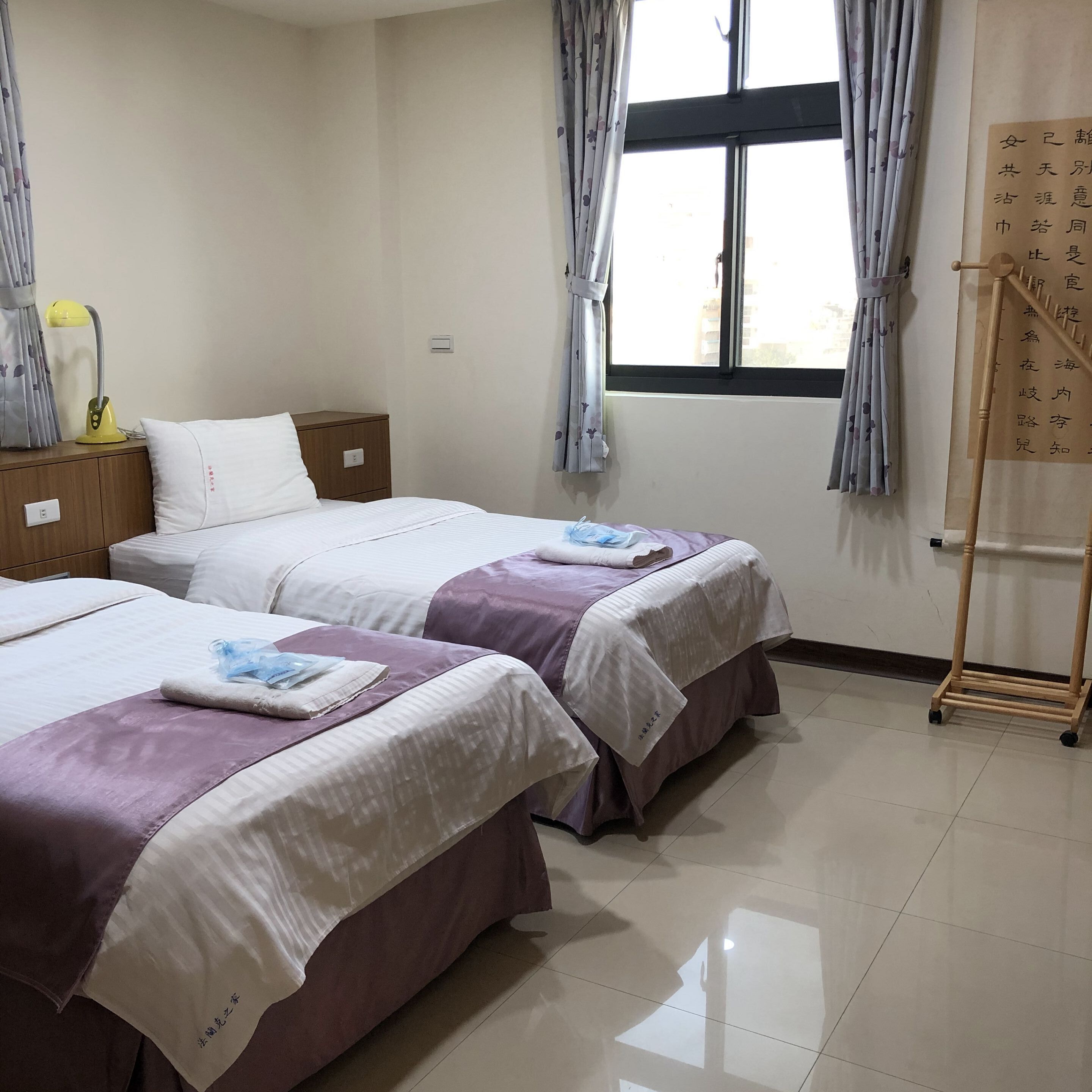 Quarto de hotel com duas camas, mesa de cabeceira com abajur, janela com cortinas e um suporte de madeira com papéis.