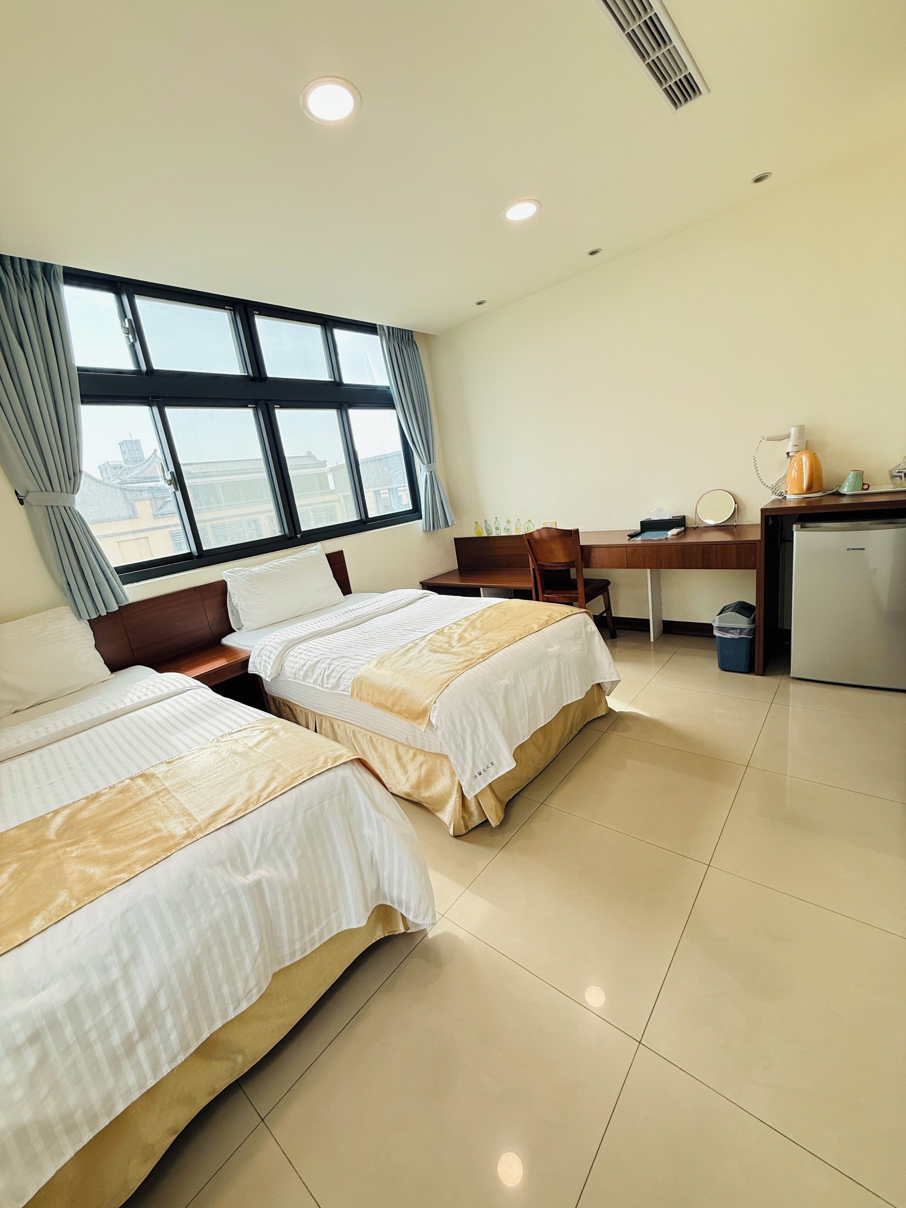 Quarto de hotel com duas camas, uma escrivaninha, uma cadeira, um mini frigorífico e uma janela com cortinas.