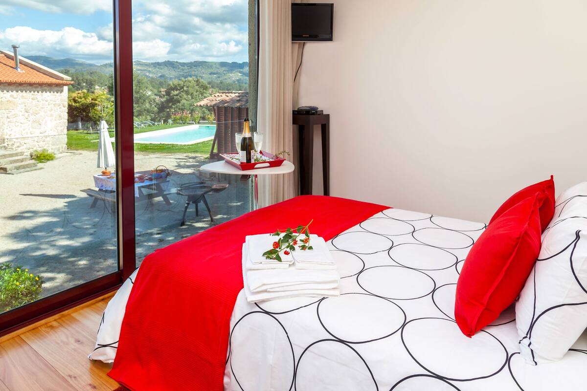 Quarto com uma grande janela, cama com colcha estampada em vermelho e branco, pequena mesa com bandeja de bebidas e vista para uma piscina e montanhas.