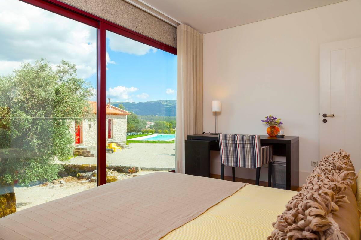 Quarto com uma cama grande, uma escrivaninha com luminária e vista para uma piscina e montanhas.