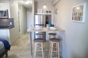 Cozinha compacta com geladeira, micro-ondas e banquetas. Há uma mesa de jantar com pratos e copos.