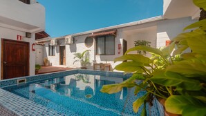 Piscina com borda azulejada, rodeada por um edifício branco com porta e janelas de madeira, e plantas em vasos.