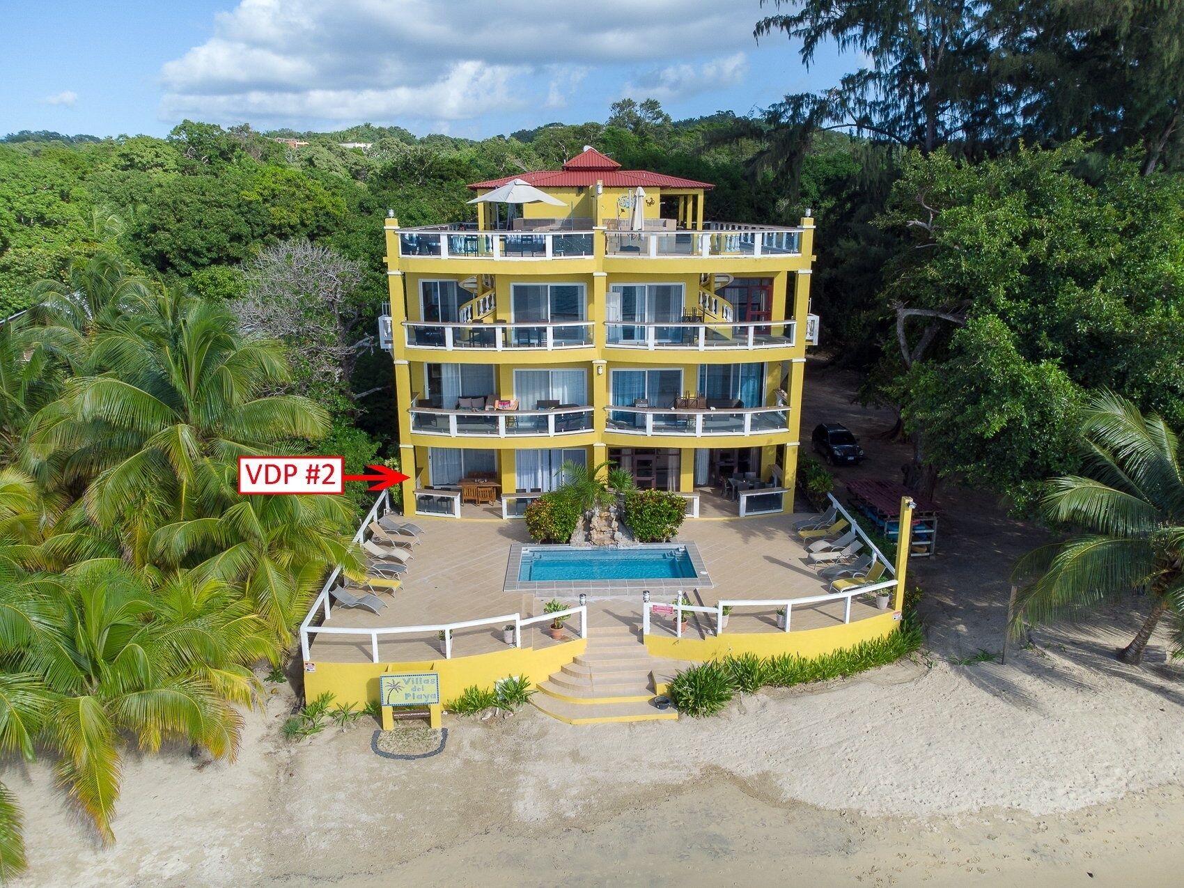 Um edifício amarelo de vários andares com piscina, cercado por palmeiras e uma praia.