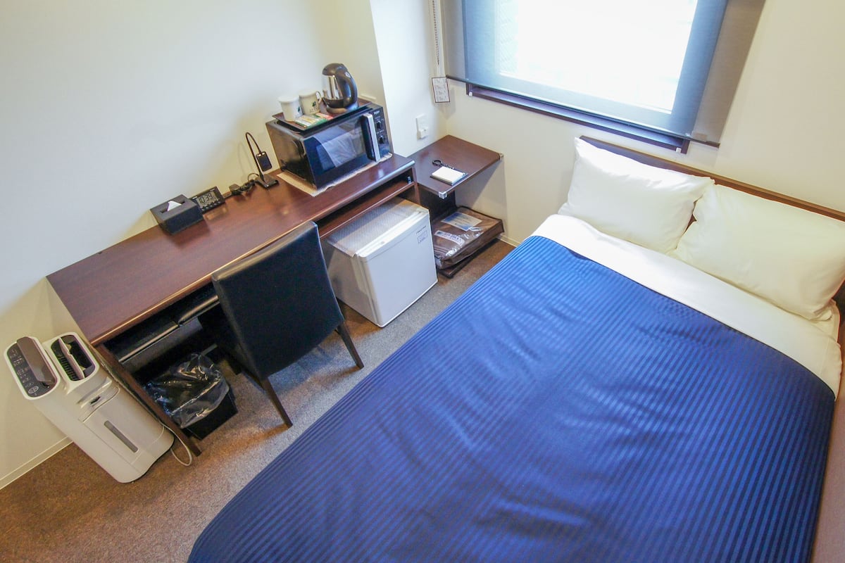 Quarto de hotel com cama, escrivaninha com computador, micro-ondas, cafeteira e pequeno frigorífico.