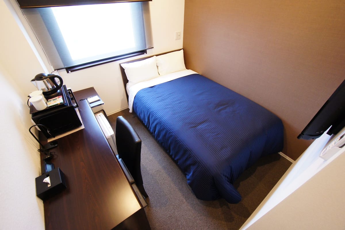 Quarto pequeno com uma cama de solteiro, escrivaninha, cadeira e janela.