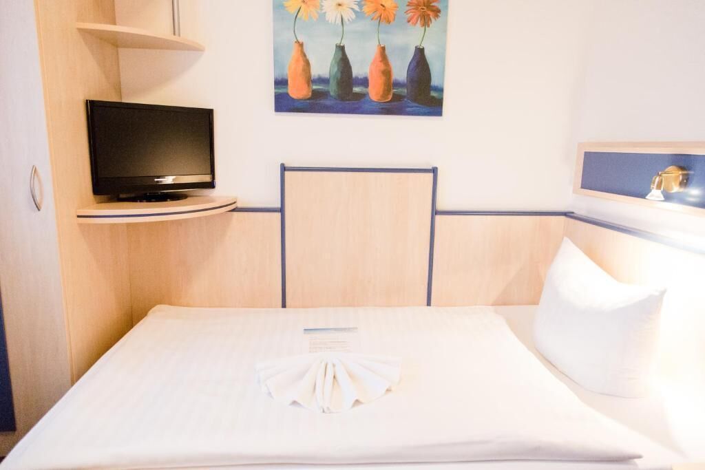 Quarto de hotel com cama, televisão sobre suporte, pintura de vasos e pequena estante com abajur.