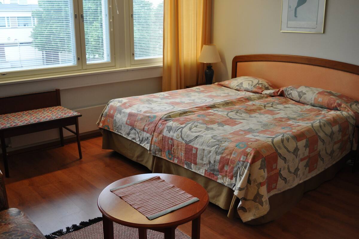 Quarto de hotel com cama, um banquinho, uma mesinha e janela com persianas.