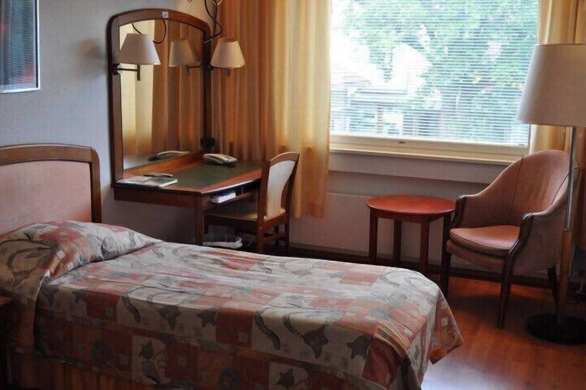 Quarto de hotel com uma cama, uma mesa com telefone, uma cadeira e uma pequena mesa.