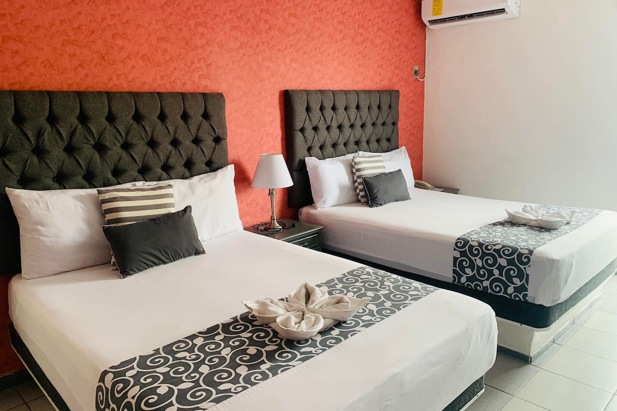 Quarto de hotel com duas camas, cada uma com cabeceira estofada, roupa de cama branca e almofadas decorativas. Há um abajur numa cama e uma toalha na outra.