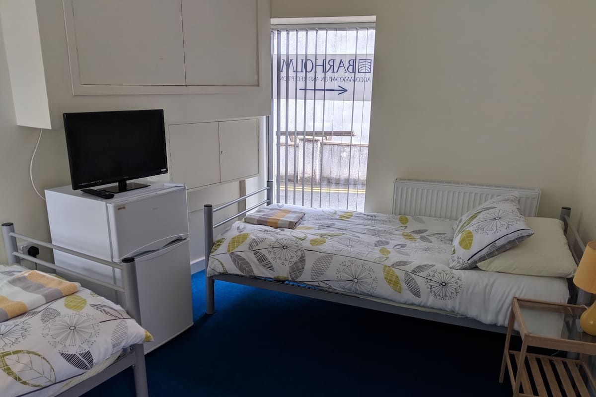 Quarto com cama, escrivaninha com computador, cadeira e janela com vista para um edifício.