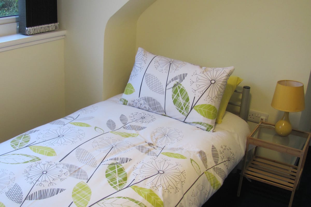 Uma cama de solteiro com uma colcha de padrão floral, uma mesinha de cabeceira com abajur e um radiador.