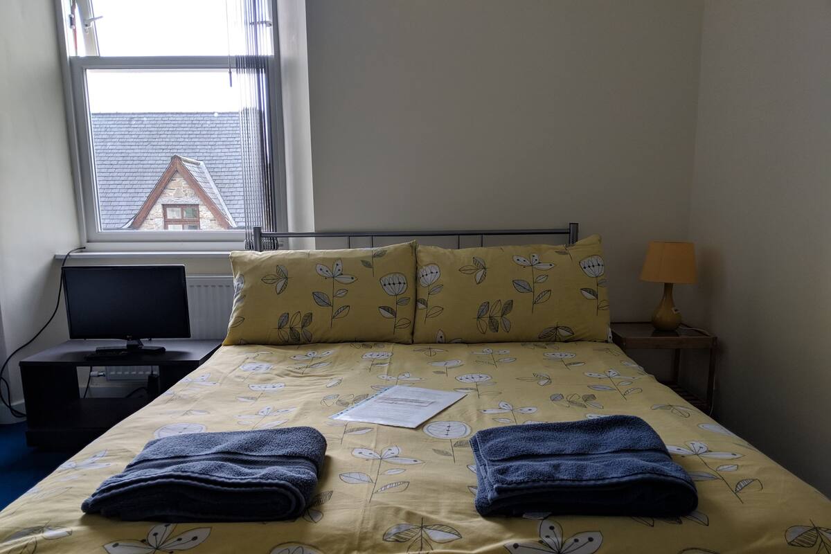 Uma cama bem arrumada com roupa de cama florida, uma luminária de cabeceira e um computador sobre uma escrivaninha.