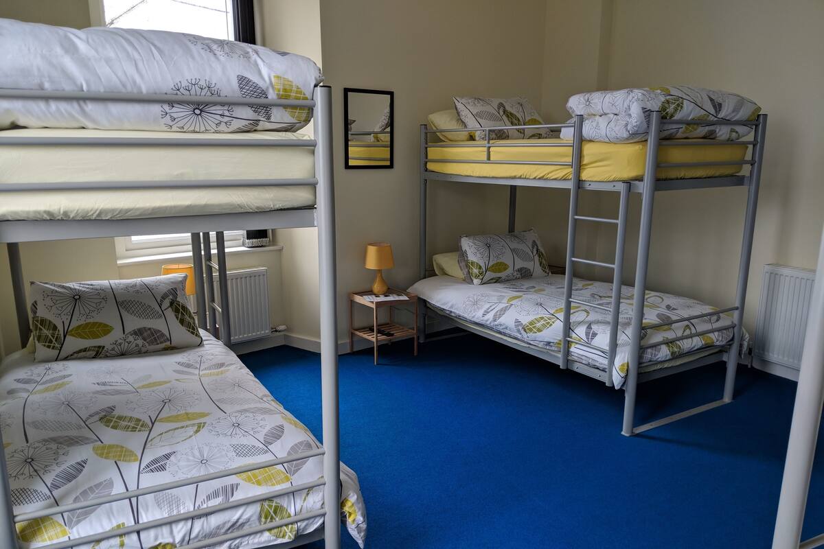 Quarto com beliche, carpete azul, uma pequena mesa com abajur e janela com persianas.