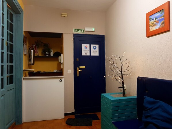 Um saguão de hotel com um balcão de recepção, uma porta azul identificada com 'SALIDA', uma pequena planta e um quadro na parede.