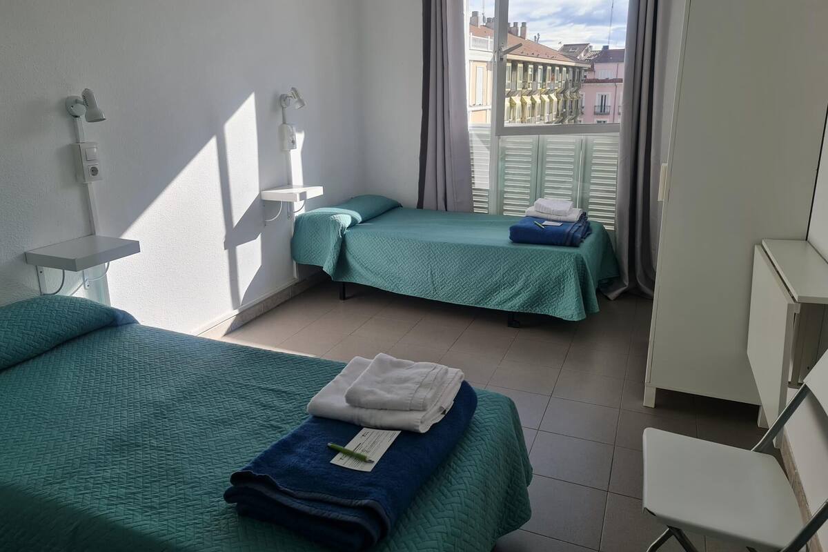 Quarto de hotel com duas camas de solteiro, uma pequena mesa de cabeceira, uma cadeira e vista para prédios pela janela.