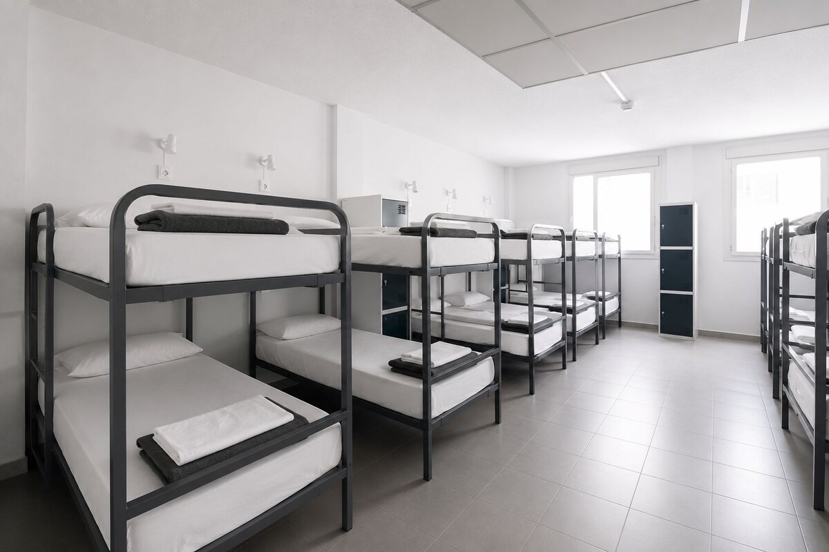 Quarto em dormitório com beliches dispostos em fileiras, uma janela e uma porta.