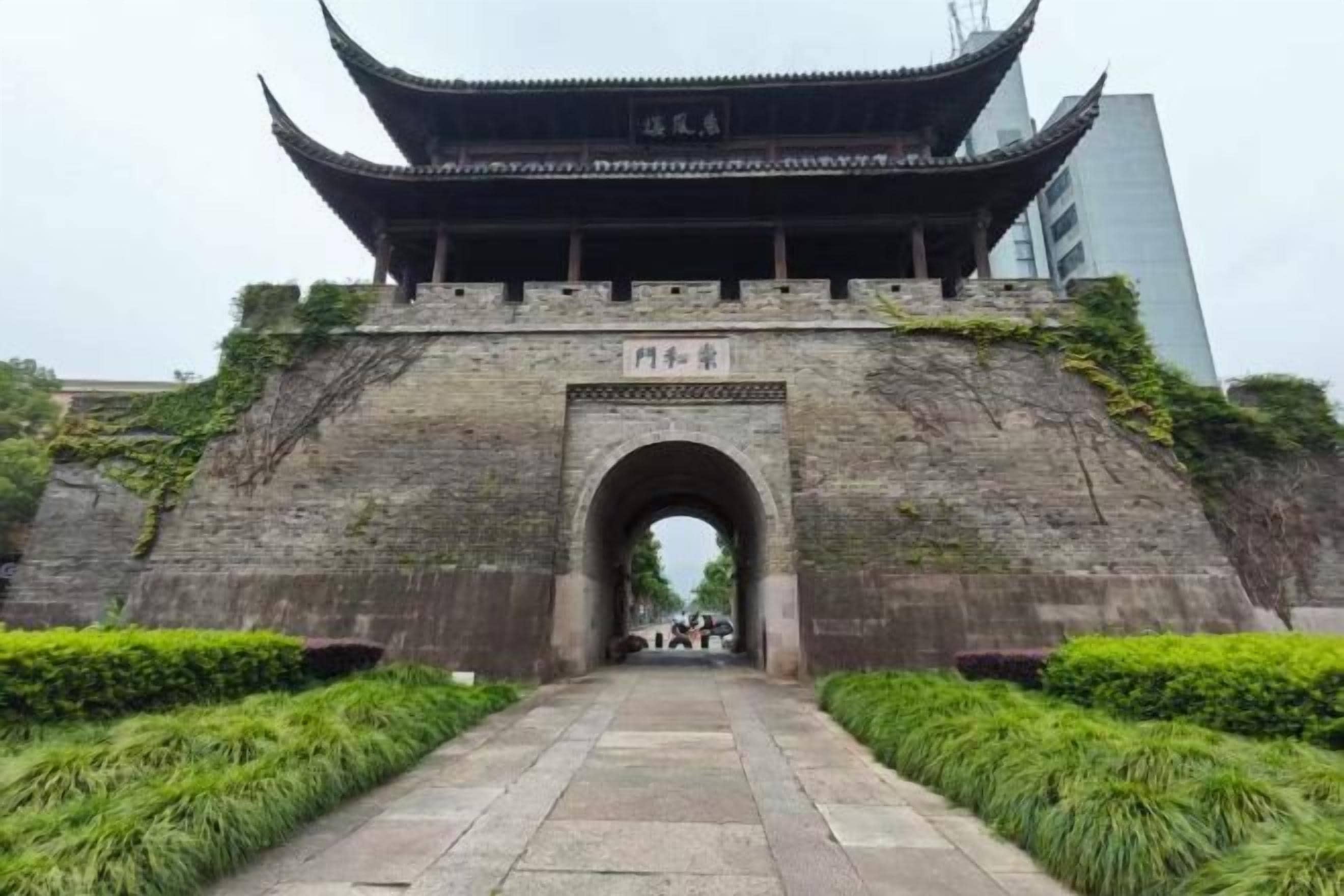 Um portão chinês tradicional com telhado ladrilhado e um arco, rodeado por vegetação.