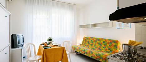 Um quarto pequeno e luminoso com sofá, mesa de jantar com cadeiras, cozinha compacta e televisão.