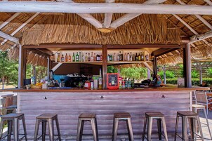 Uma área de bar ao ar livre com telhado de palha, balcão de madeira e bancos.