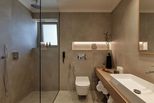 Uma casa de banho moderna com chuveiro em cabine de vidro, sanita, lavatório com espelho e prateleira com artigos de higiene pessoal.