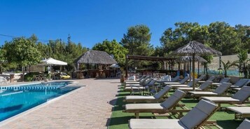 Área de piscina com espreguiçadeiras, bar e uma estrutura com telhado de palha.