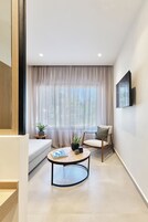Uma sala de estar moderna com sofá, mesa de centro redonda, poltrona, TV fixada na parede e uma janela grande com cortinas leves.