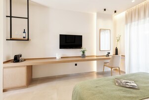 Quarto de hotel moderno com televisão de tela plana na parede, escrivaninha de madeira com cafeteira, uma cadeira e um banquinho verde com um livro.