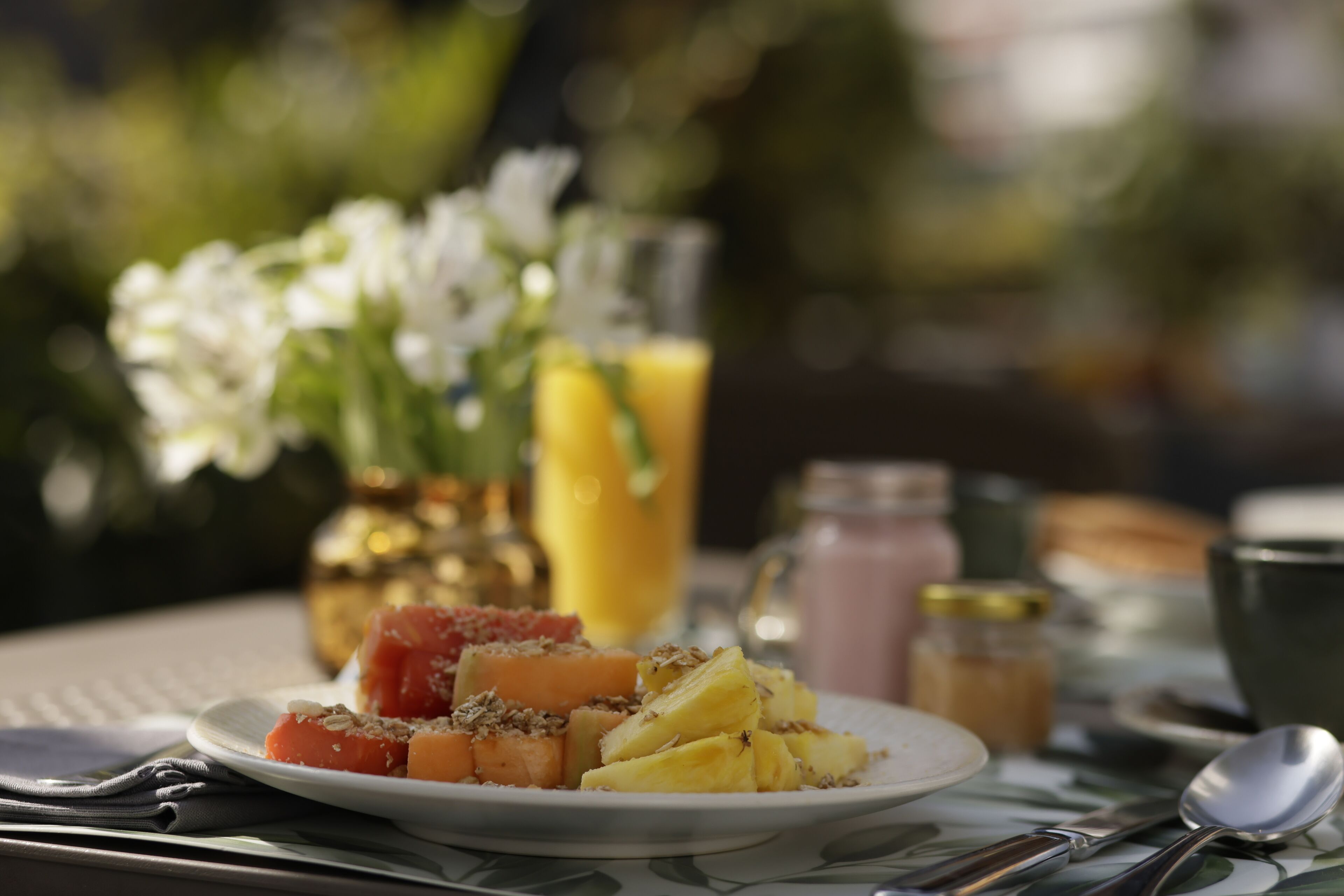 Mesa de café da manhã com uma travessa de frutas, um copo de suco de laranja e um vaso de flores.