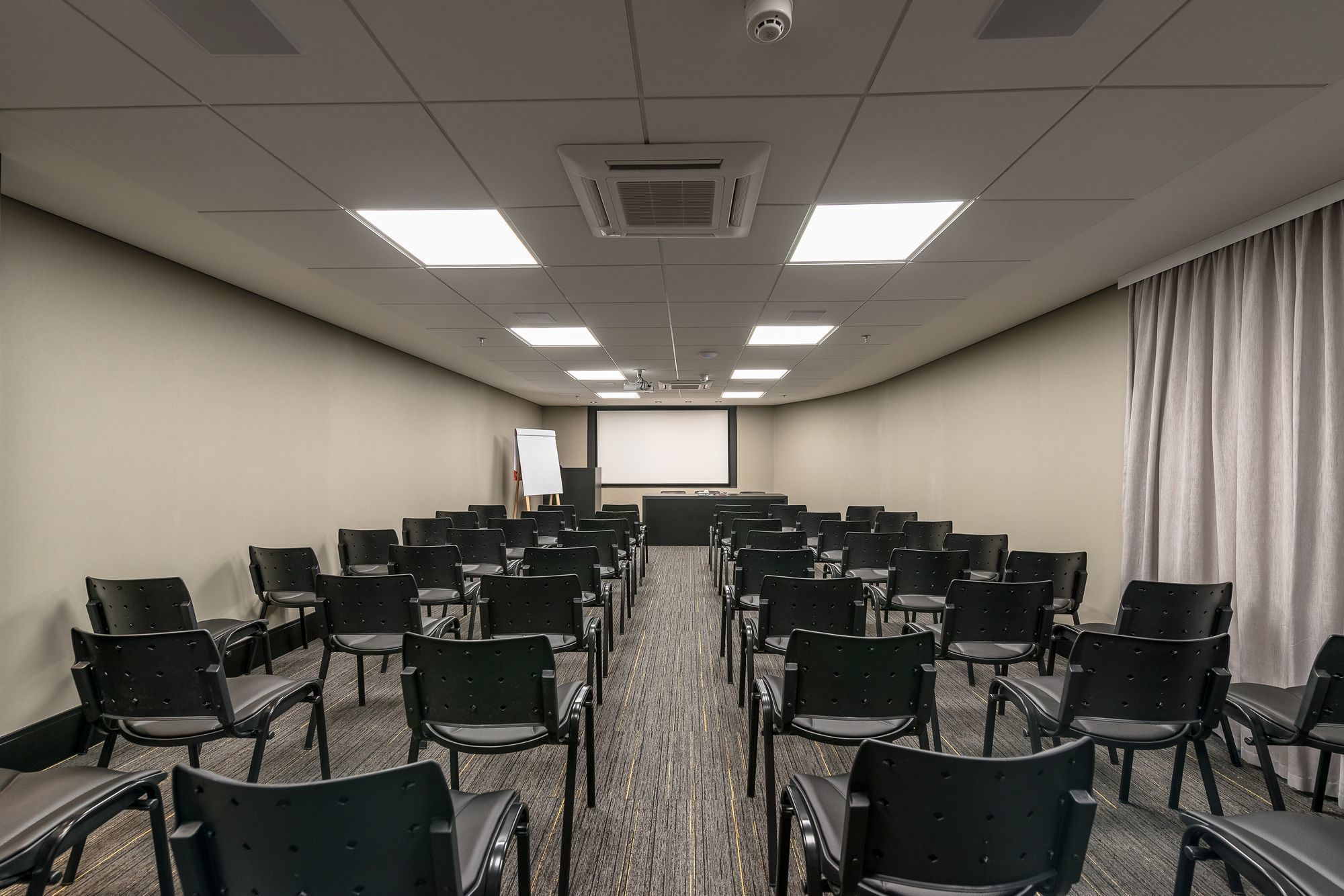 Uma sala de conferência com fileiras de cadeiras pretas, uma tela de projetor e um quadro branco.