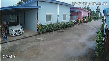 Um bairro residencial com uma casa azul, um carro branco estacionado na entrada e uma casa rosa ao fundo.