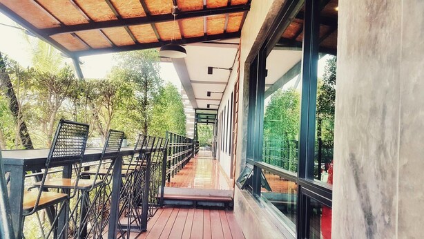 Uma área externa coberta com deck de madeira, cadeiras de metal e guarda-corpo de vidro com vista para o verde.