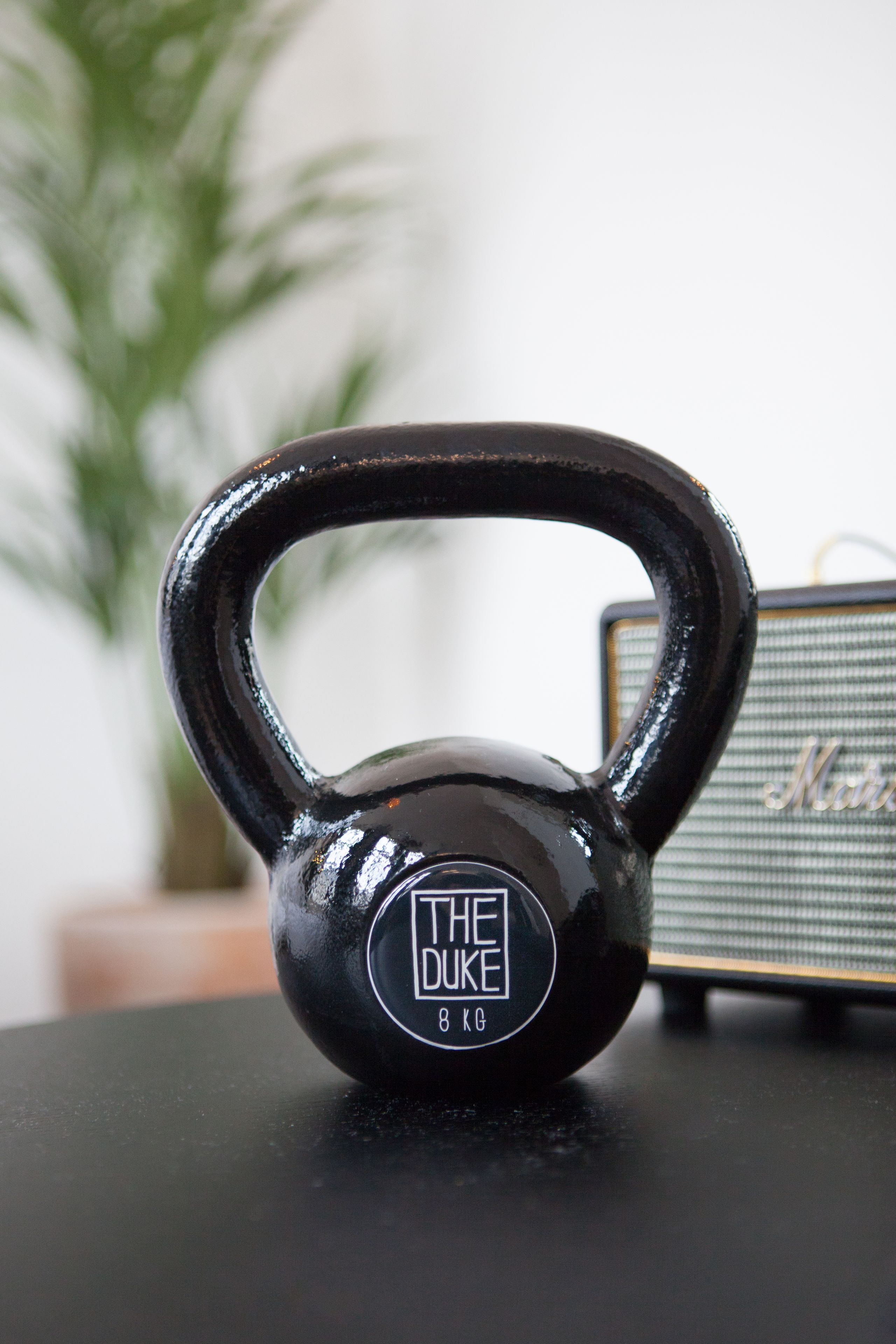 Um kettlebell preto com a inscrição "THE DUKE" e "8 KG", apoiado em uma superfície preta com fundo desfocado.