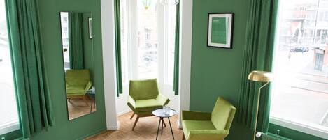 Quarto com duas poltronas verdes, uma mesinha redonda, abajur, espelho e janela com vista para prédios.