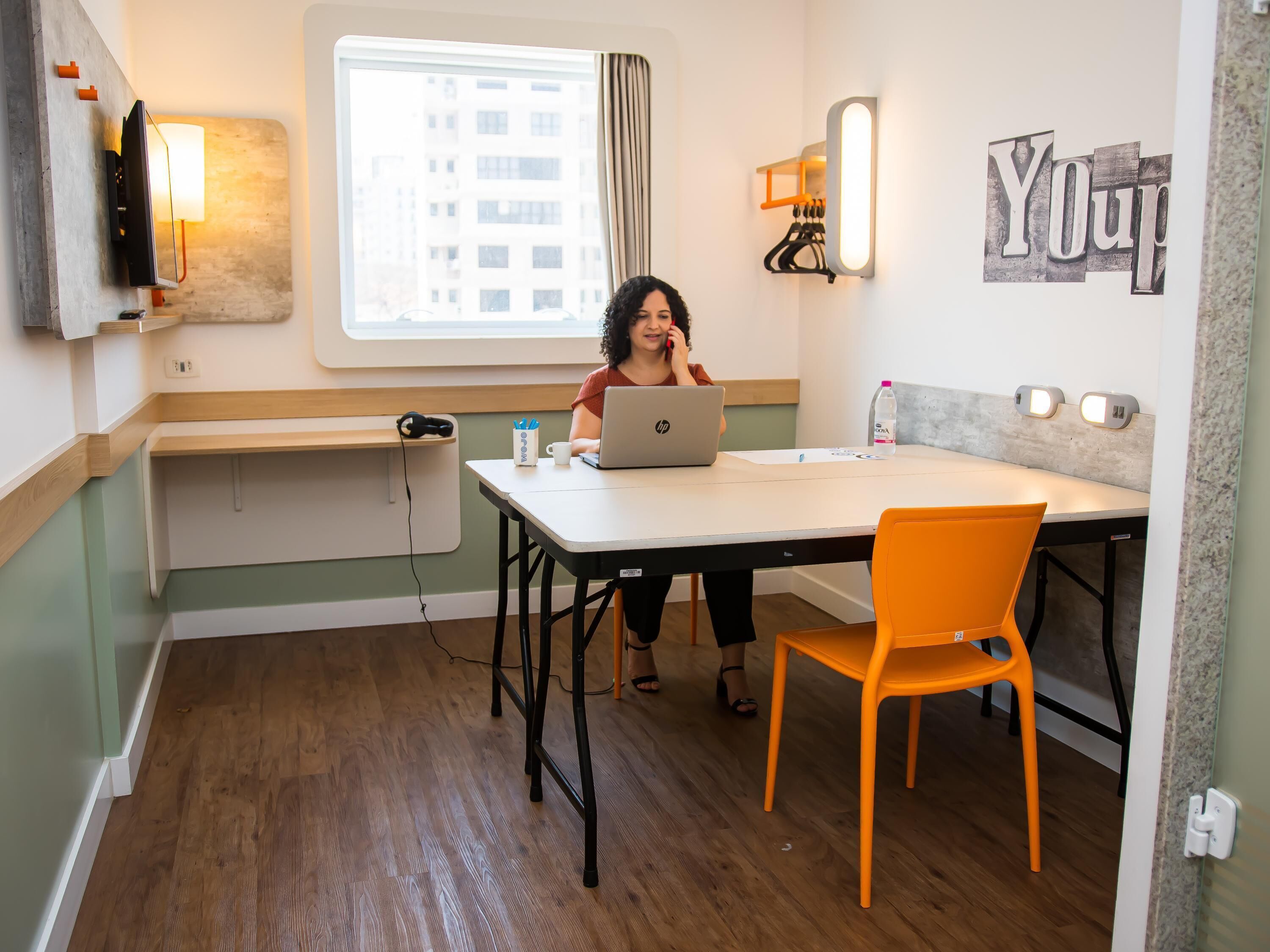 Quarto com escrivaninha, cadeira, laptop e janela com vista para edifícios.
