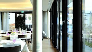 Uma área de refeições moderna, com mesas preparadas para o serviço, grandes janelas com vista para o exterior e um design interior limpo e minimalista.