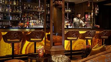 Uma área de bar com várias prateleiras de garrafas de bebidas, um bartender preparando uma bebida, e assentos com um sofá e poltronas confortáveis.