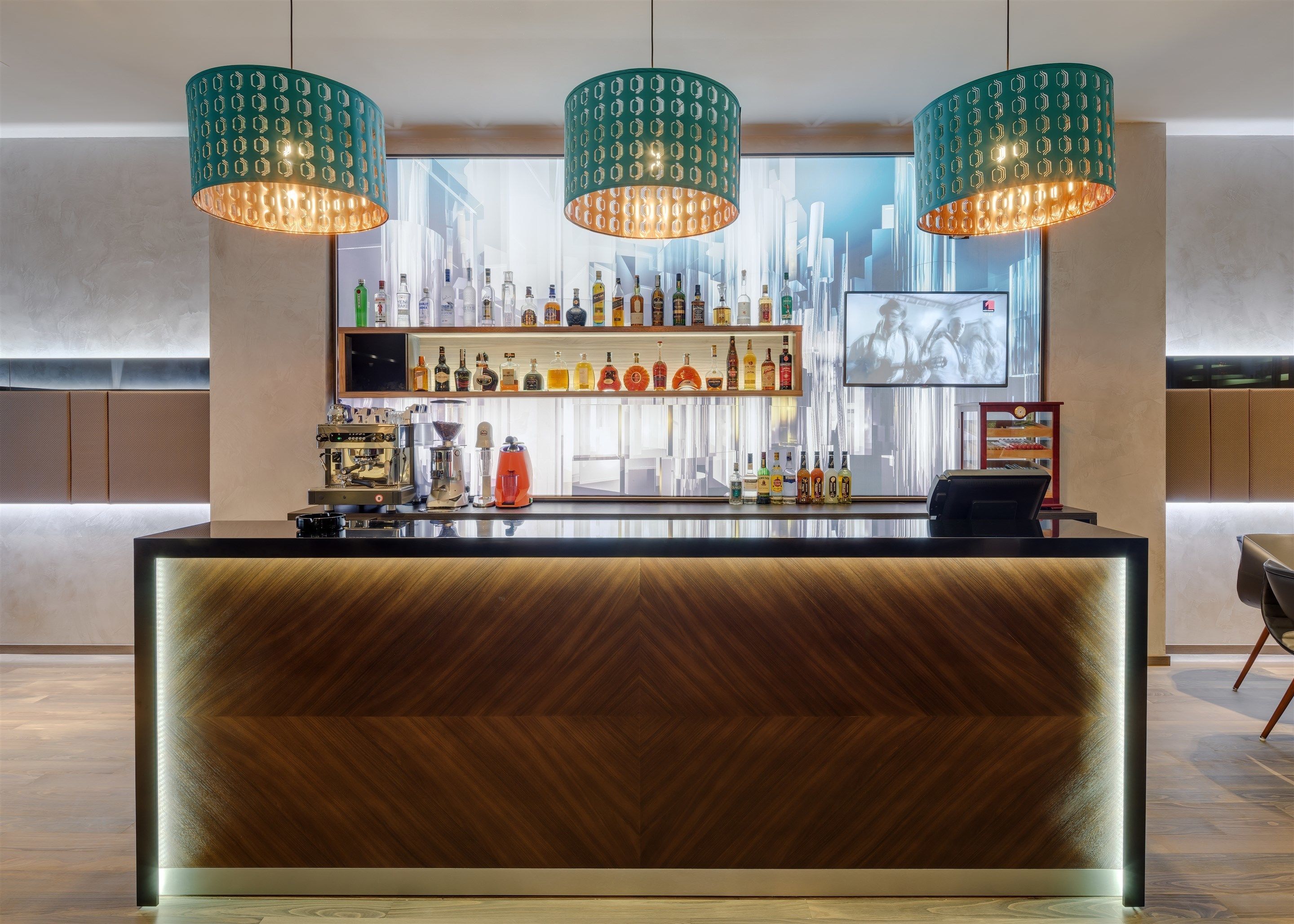 Uma área de bar moderna com balcão de madeira, três luminárias pendentes hexagonais e um mural com vista para a cidade ao fundo.
